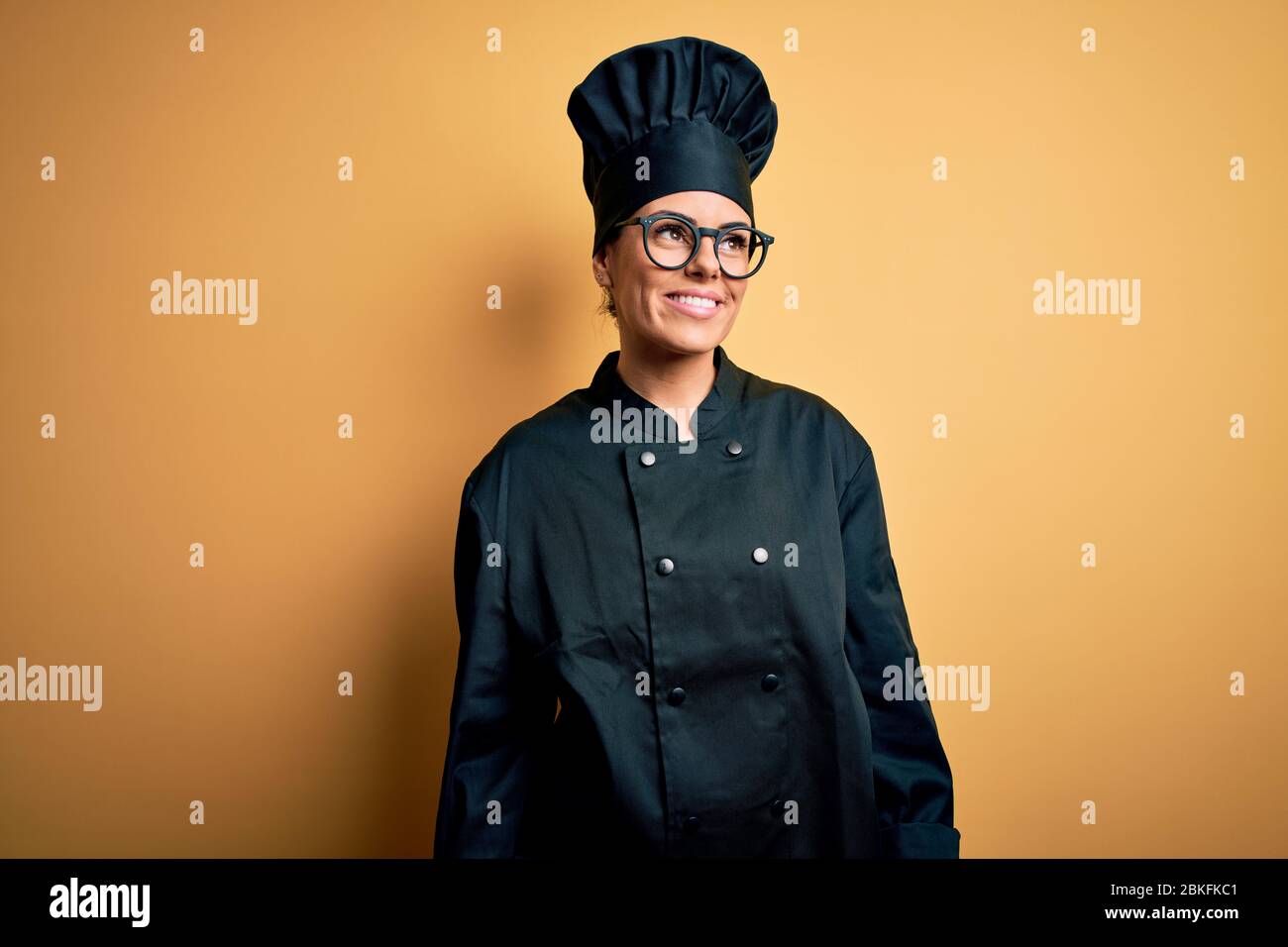 Young beautiful brunette chef woman wearing cooker uniform and hat over ...