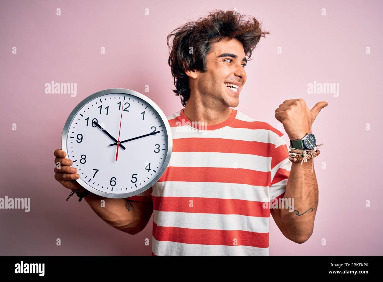 Young handsome man holding big clock standing over isolated pink ...
