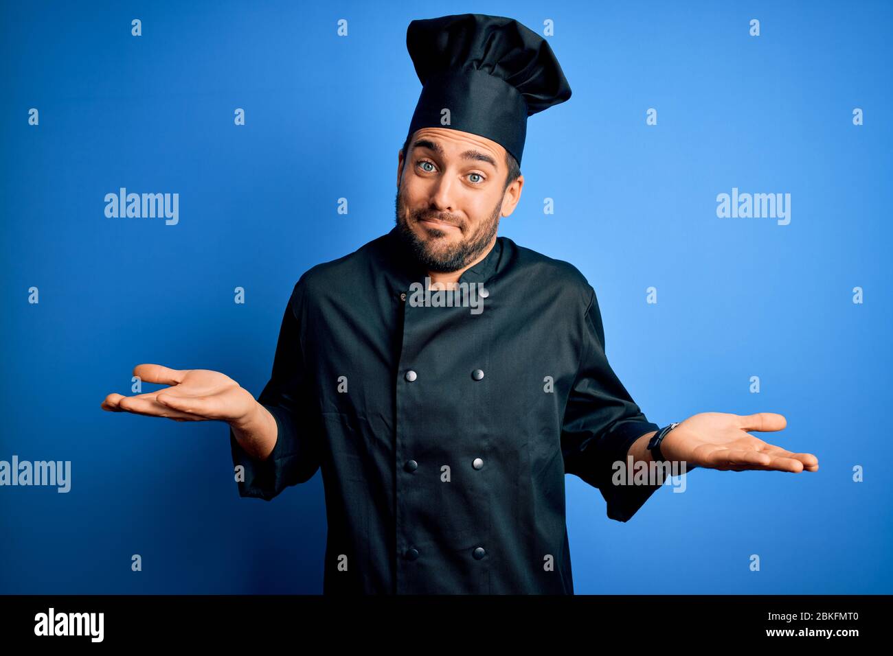 Young handsome chef man with beard wearing cooker uniform and hat over ...