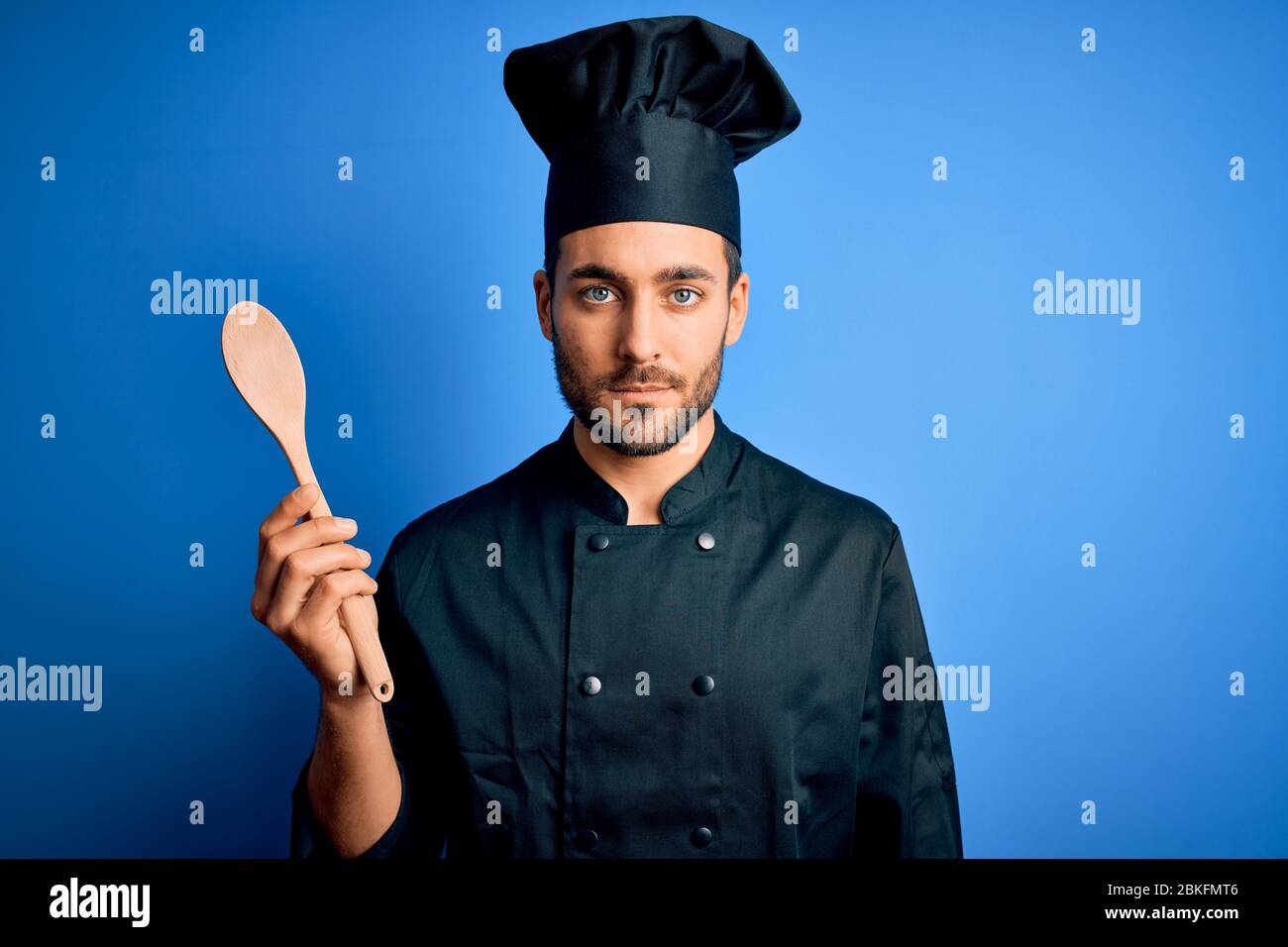 Young cooker man with beard wearing uniform holding wooden spoon over ...