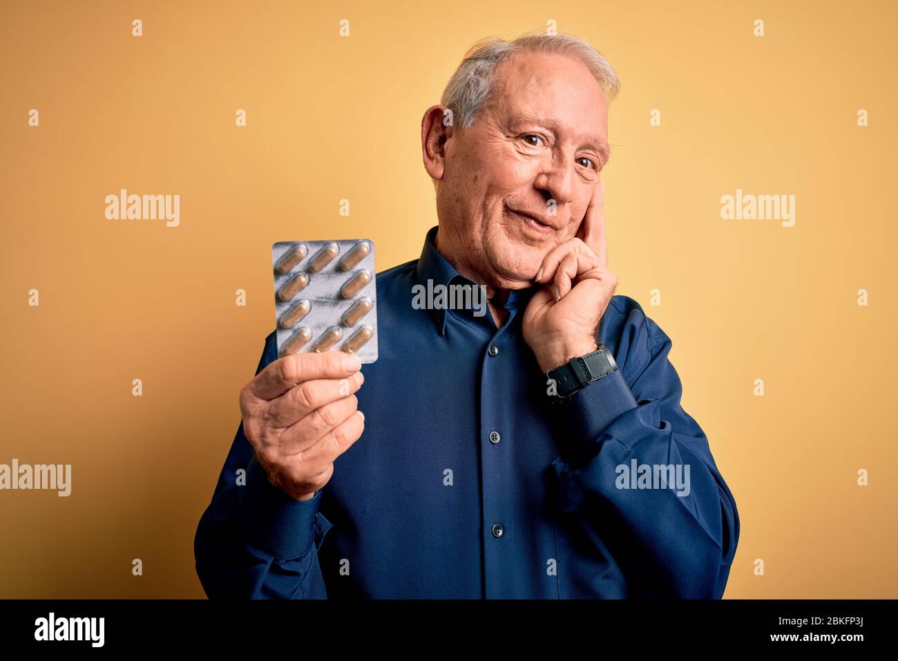 Senior grey haired man holding pharmaceutical pill drugs over yellow ...