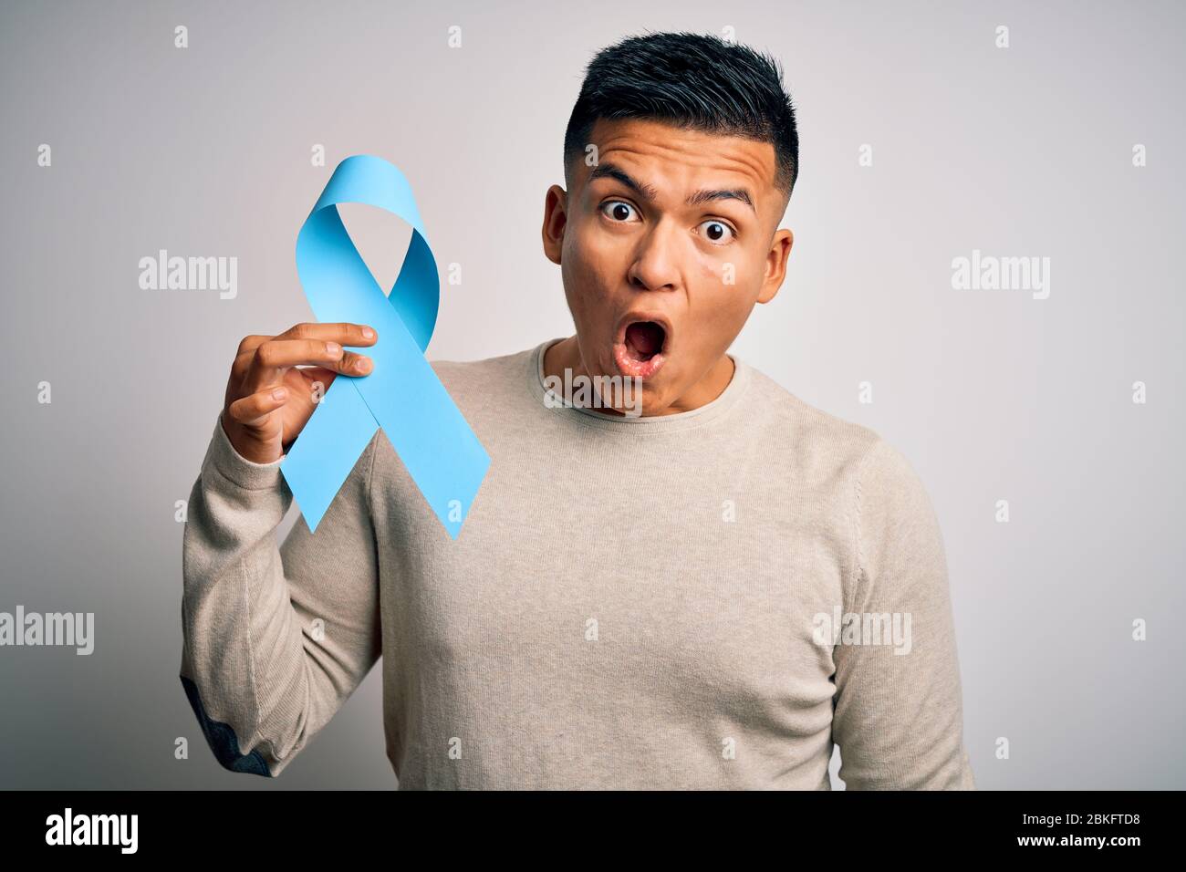 Young handsome latin man holding blue cancer ribbon over isolated white ...