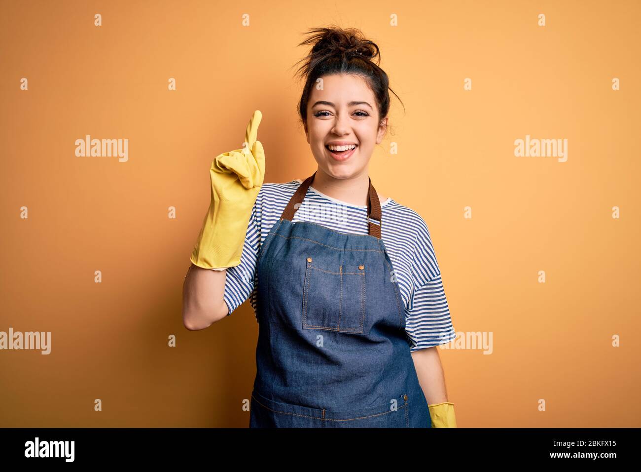 Young brunette cleaner woman wearing housekeeping gloves over yellow ...