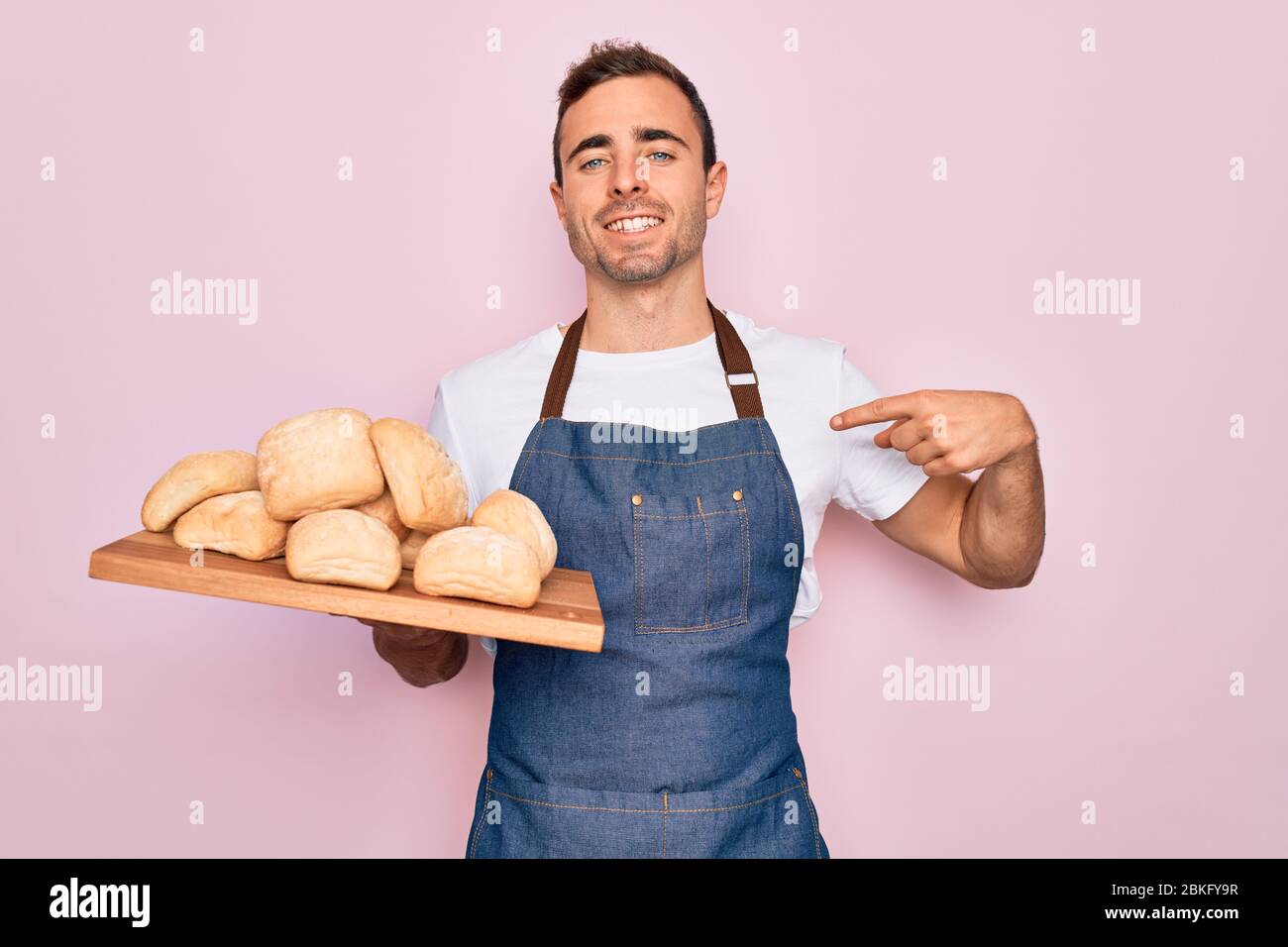 Young handsome baker man with blue eyes wearing apron holding tray with ...