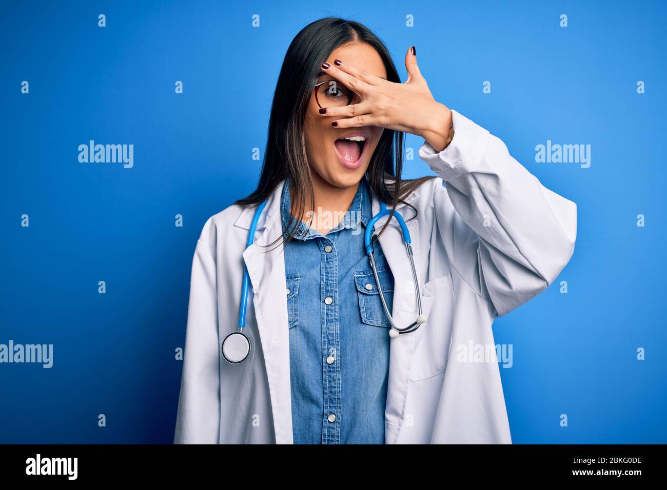 Young beautiful doctor woman wearing stethoscope and glasses over blue background peeking in ...
