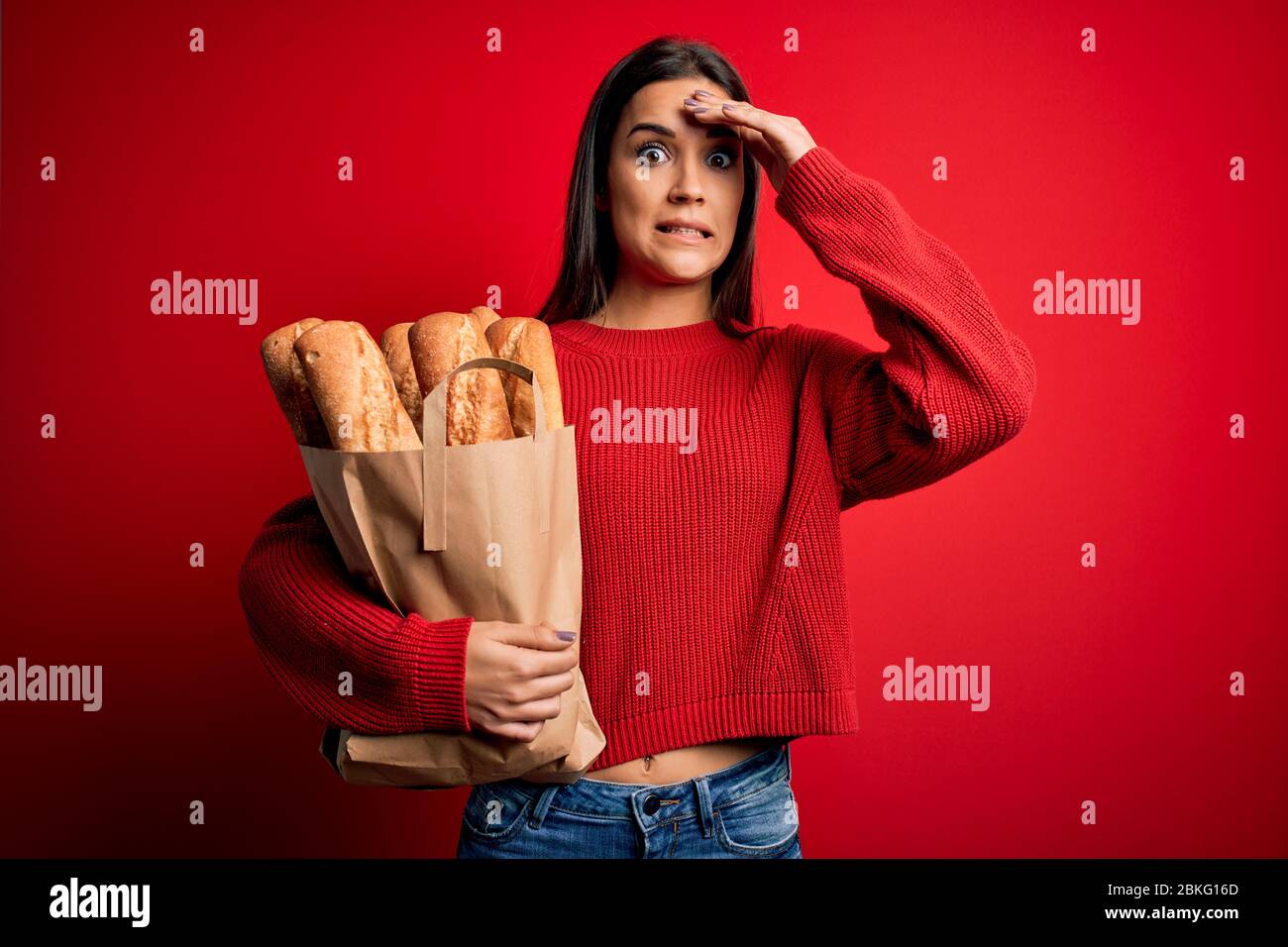 Young beautiful brunette woman holding paper bag with bread over red ...