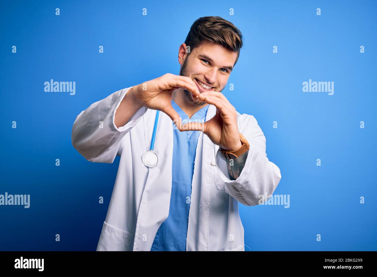 Young blond doctor man with beard and blue eyes wearing white coat and stethoscope smiling in ...