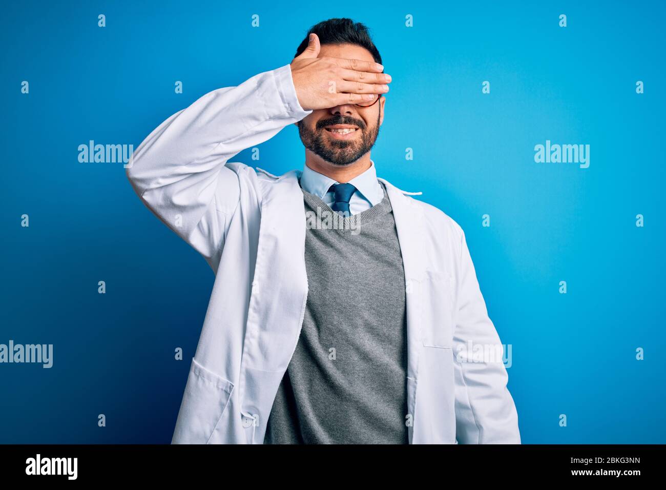 Young handsome doctor man with beard wearing coat and glasses over blue ...