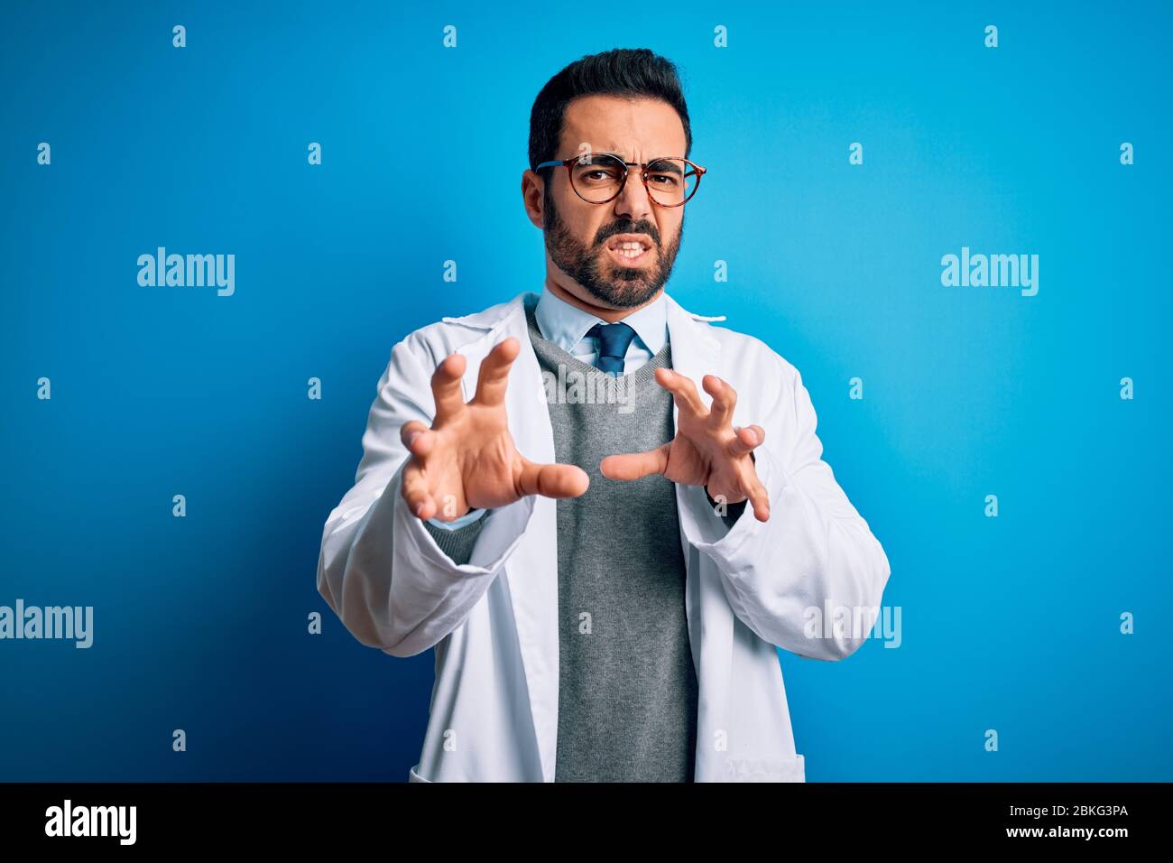 Young handsome doctor man with beard wearing coat and glasses over blue ...
