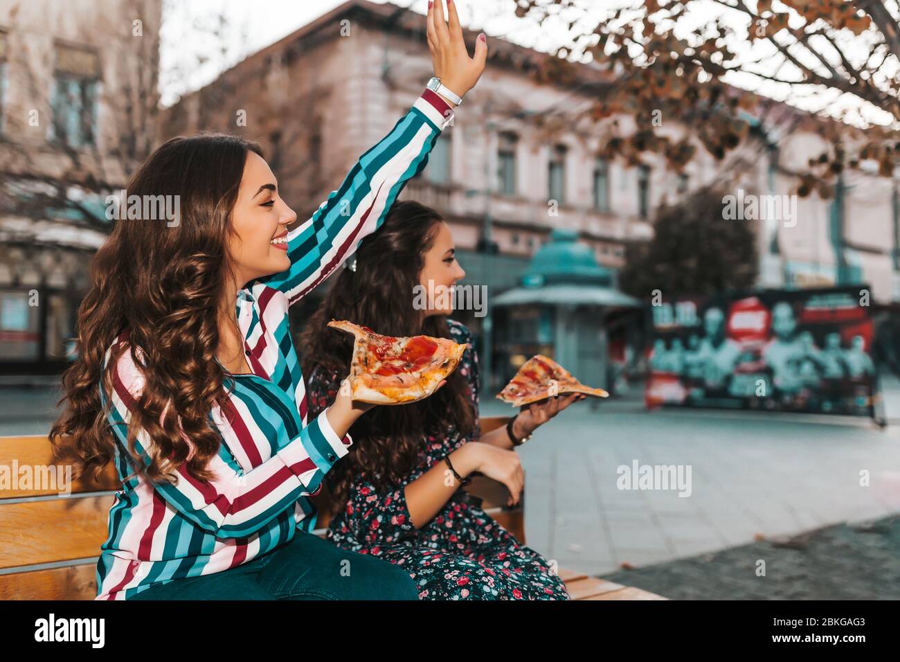 Two beautiful young women eating pizza outdoors and waving their ...