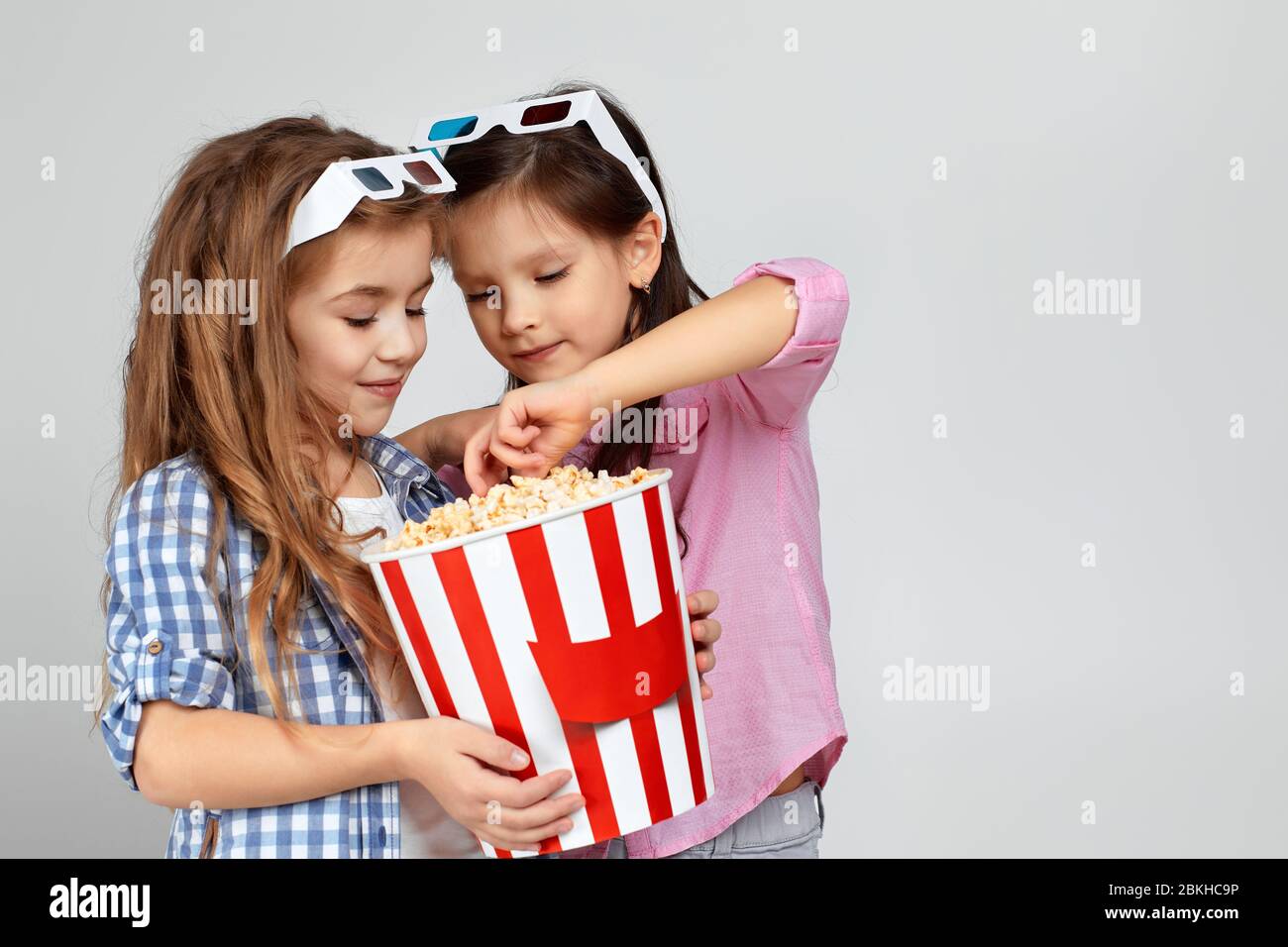 two beautiful caucasian little girls wearing red-blue 3d glasses and eating popcorn from bucket ...