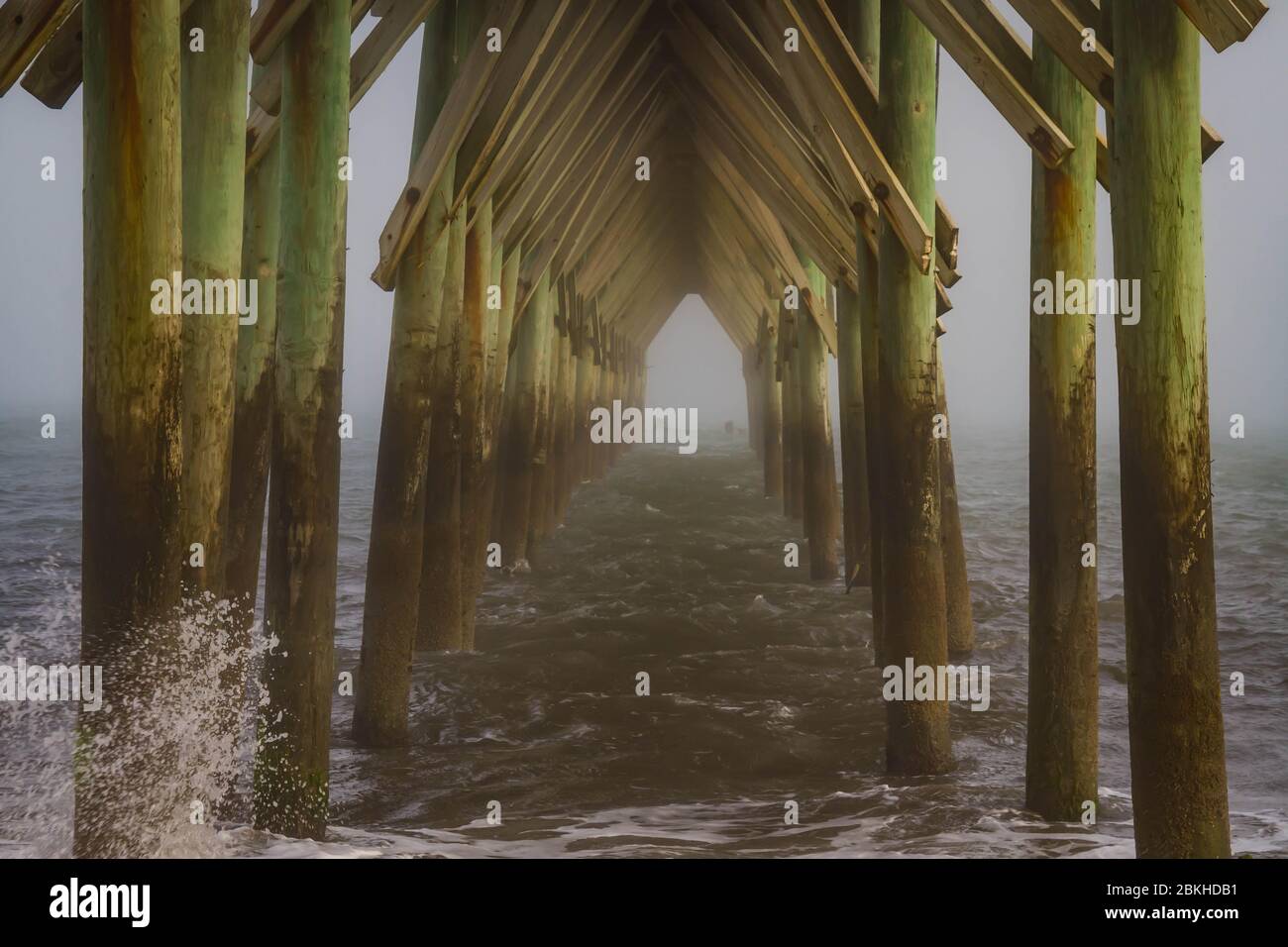 A view under a pier at a beach with rough seas and some light fog as ...