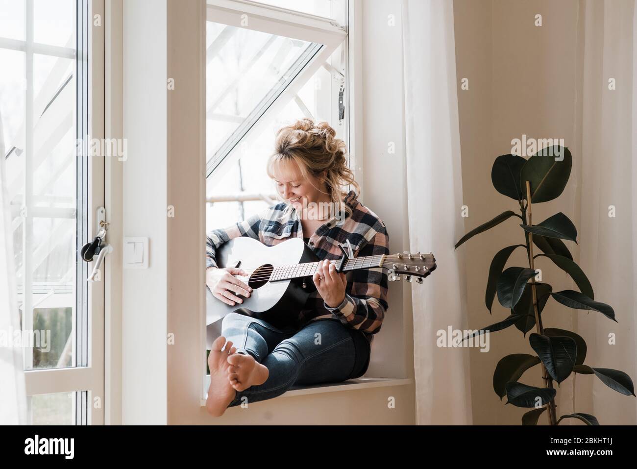woman sat at home on a window ledge smiling playing guitar Stock Photo ...