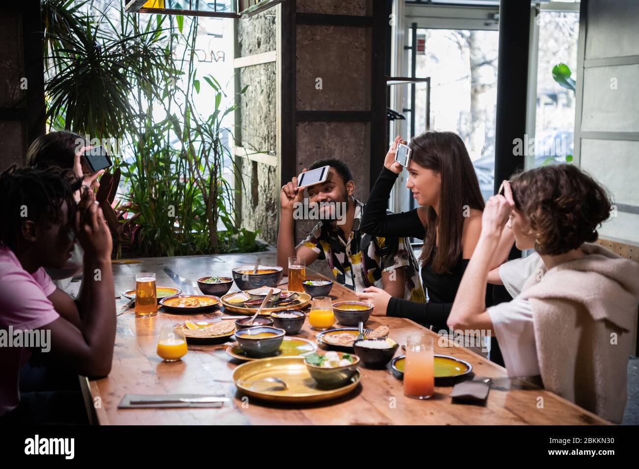 Positive Friends With Their Smartphones On Their Foreheads Sitting At The Table And Trying To positive-friends-with-their-smartphones-on-their-foreheads-sitting-at-the-table-and-trying-to