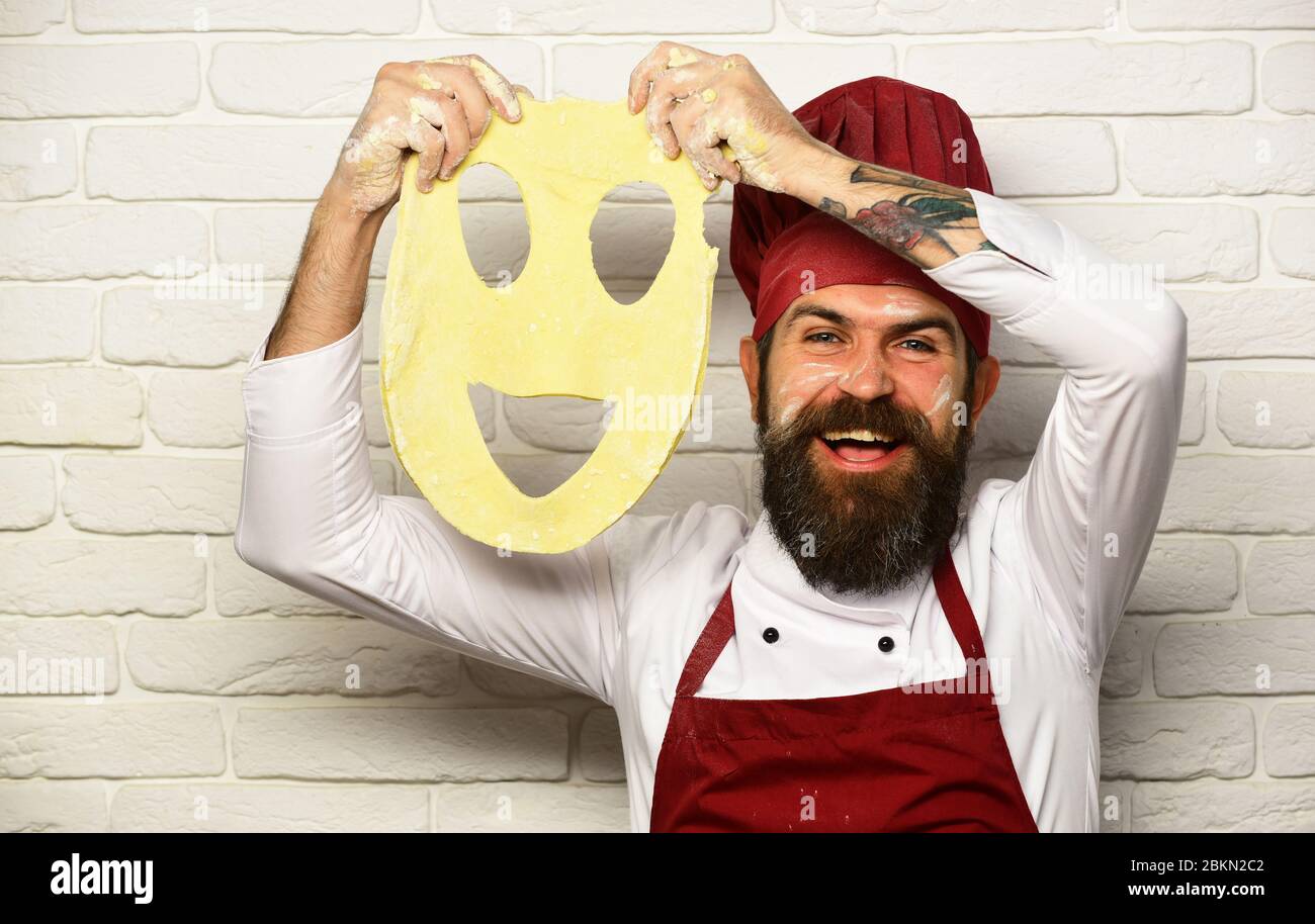 Man with beard holds rolled dough with cut smiley on white brick wall ...