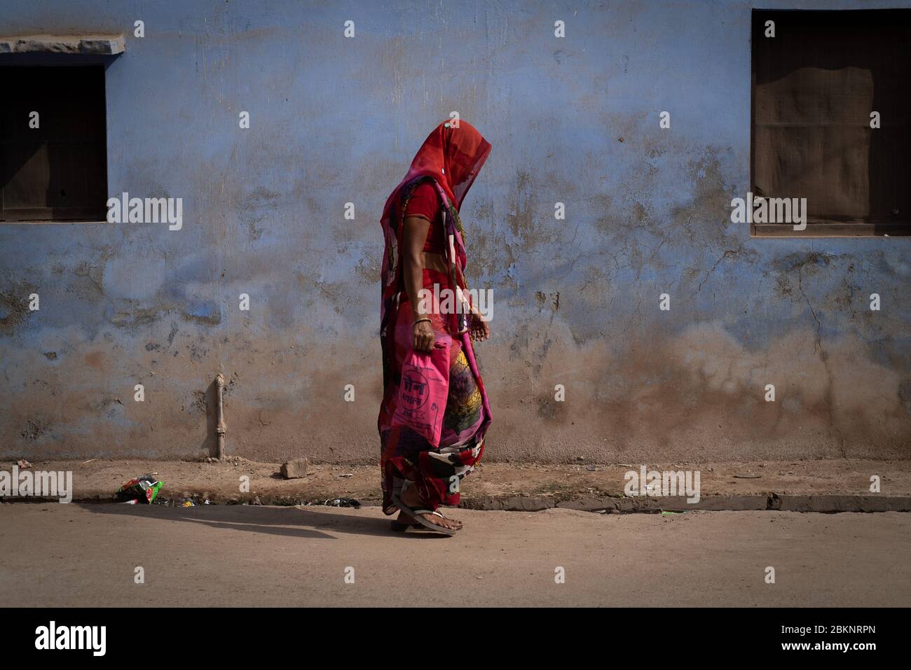 Indian woman walking, wearing a red sai against a rustic blue wall ...