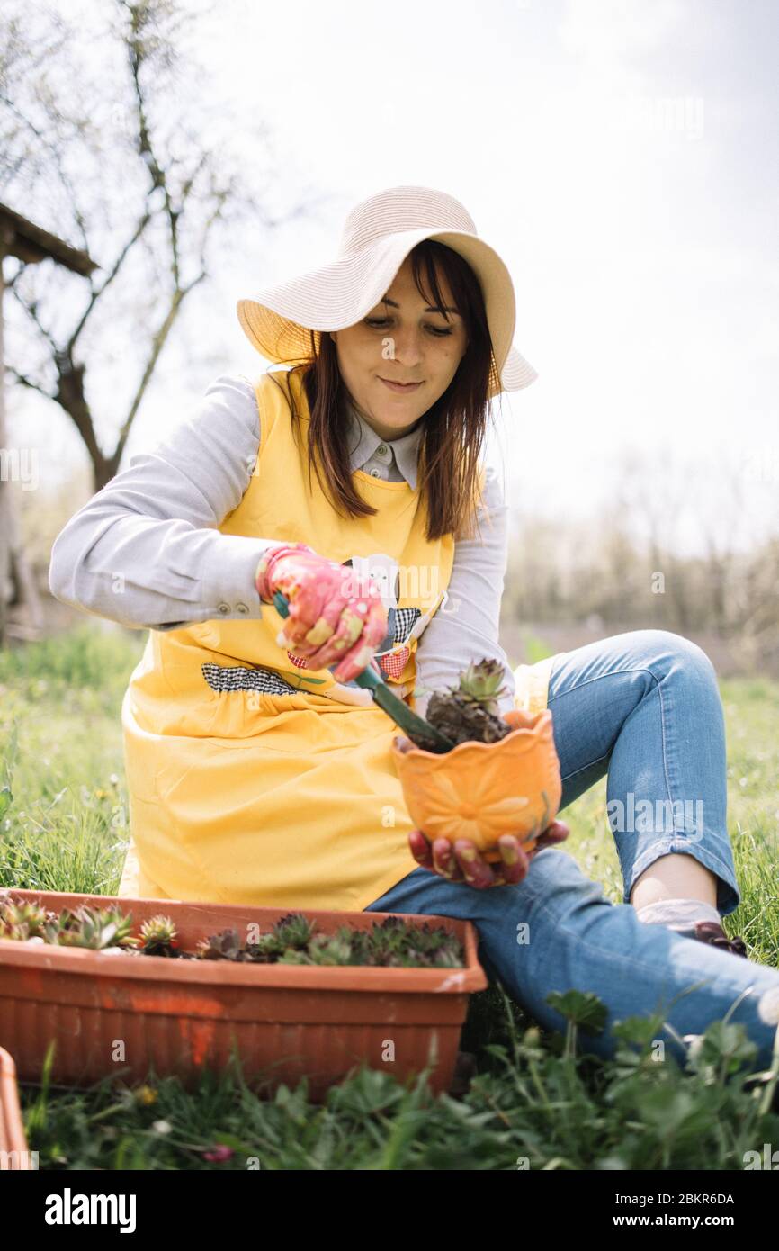 Girl putting plant into pot using hand shovel. Young woman with sun hat ...