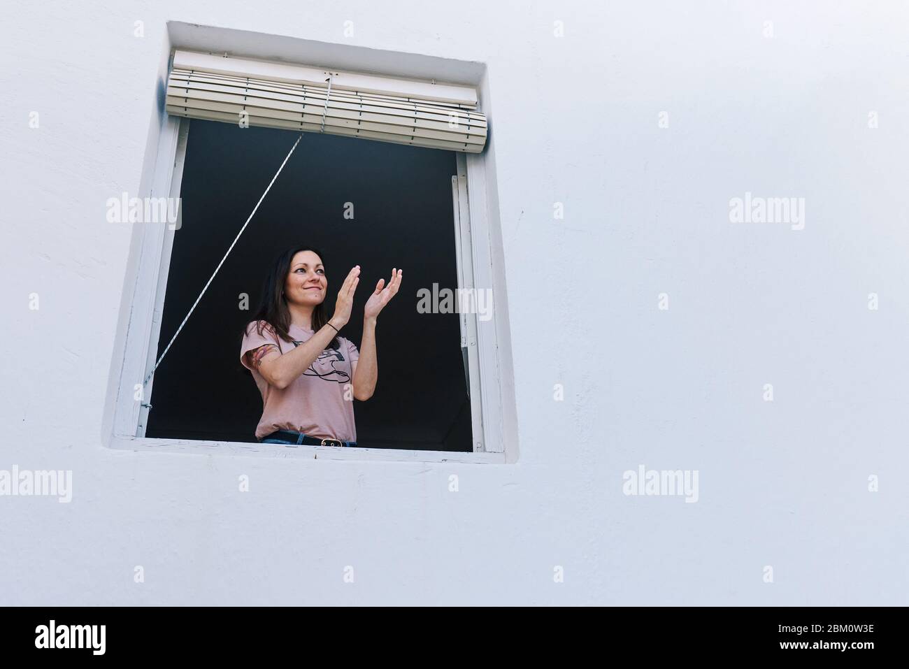 young brunette woman with mask clapping from the window of the house ...
