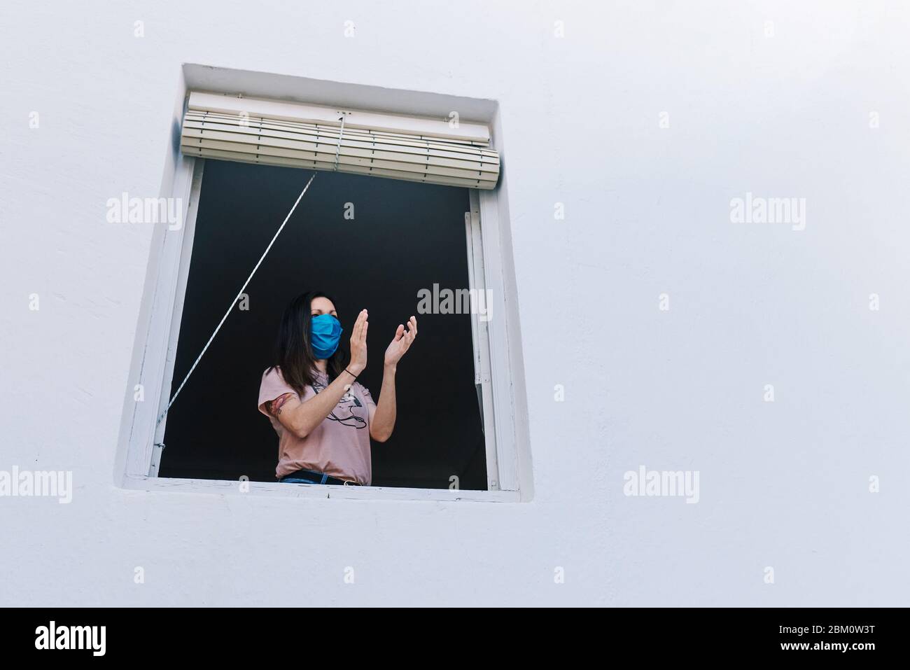 young brunette woman with mask clapping from the window of the house ...