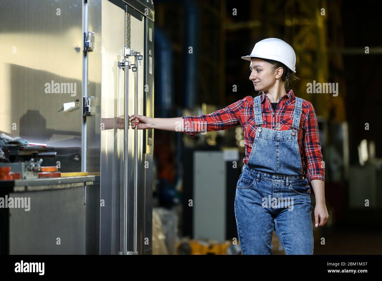 Young girl in a work dress and white hard hat in a factory. Woman in a ...