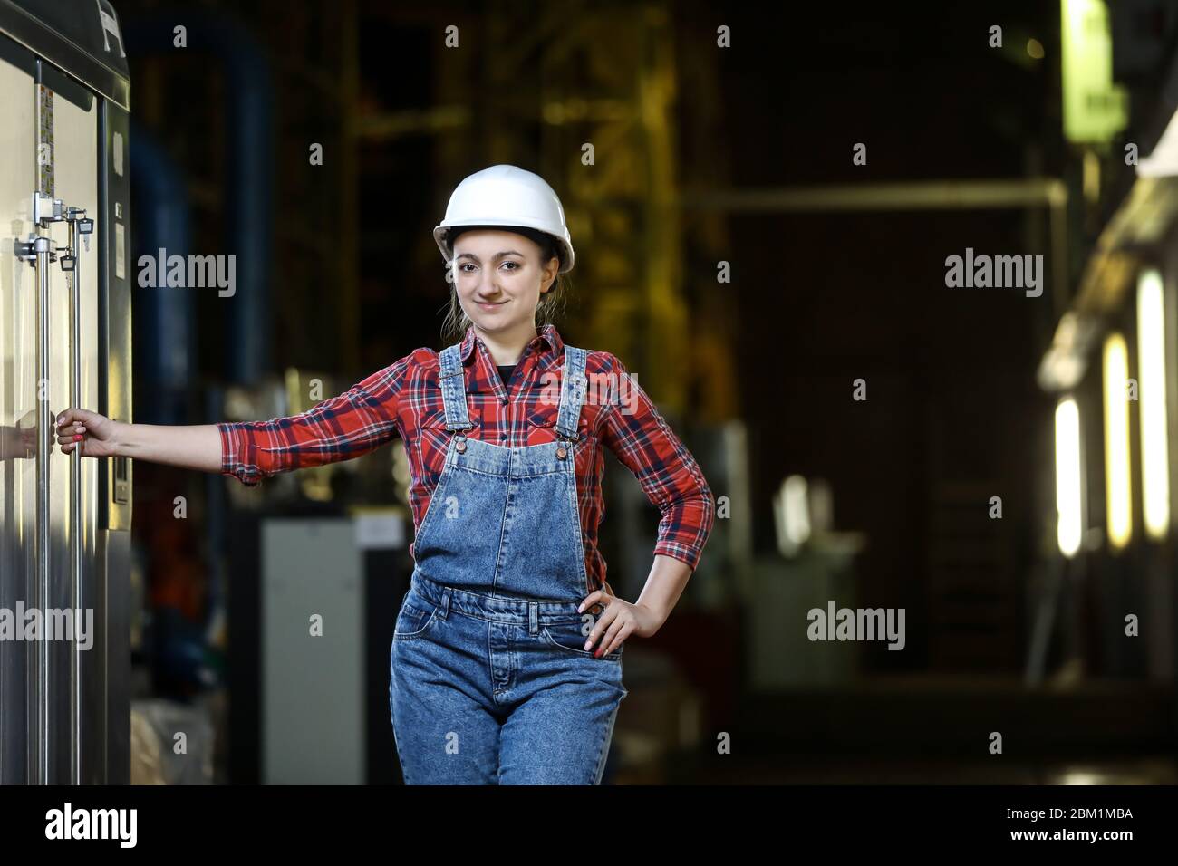 Young girl in a work dress and white hard hat in a factory. Woman in a ...