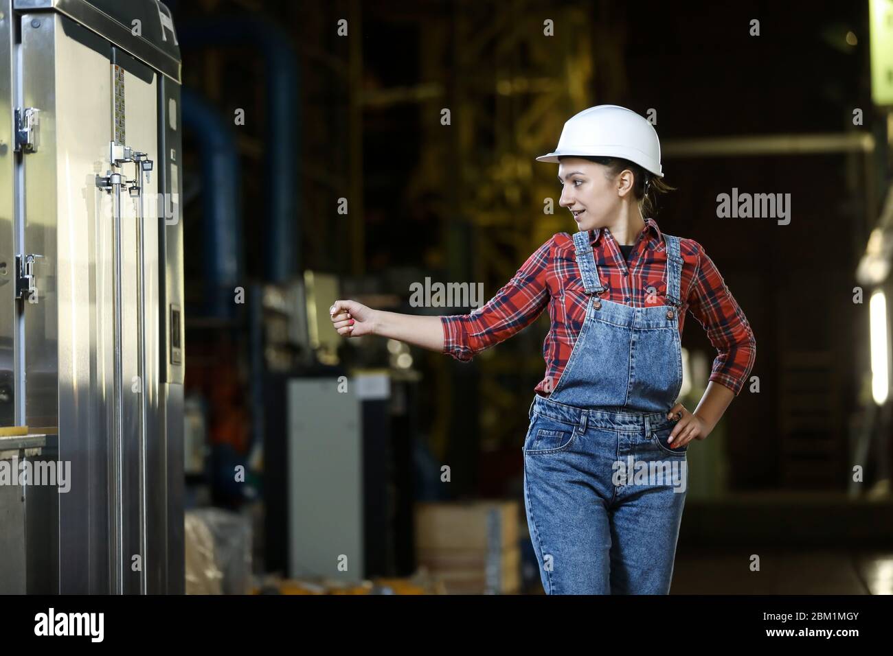 Young girl in a work dress and white hard hat in a factory. Woman in a ...