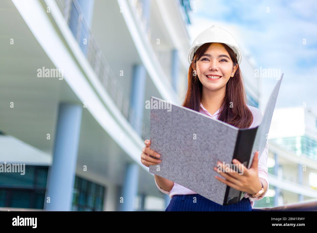 Happy young attractive female Asian engineer wearing hard hat holding ...