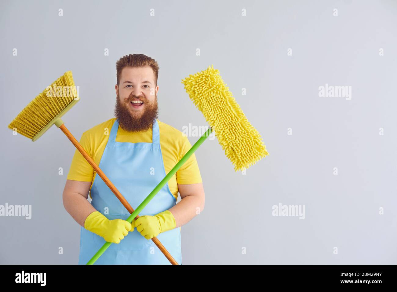 Funny fat man with a beard in an apron cleans up on a gray background ...