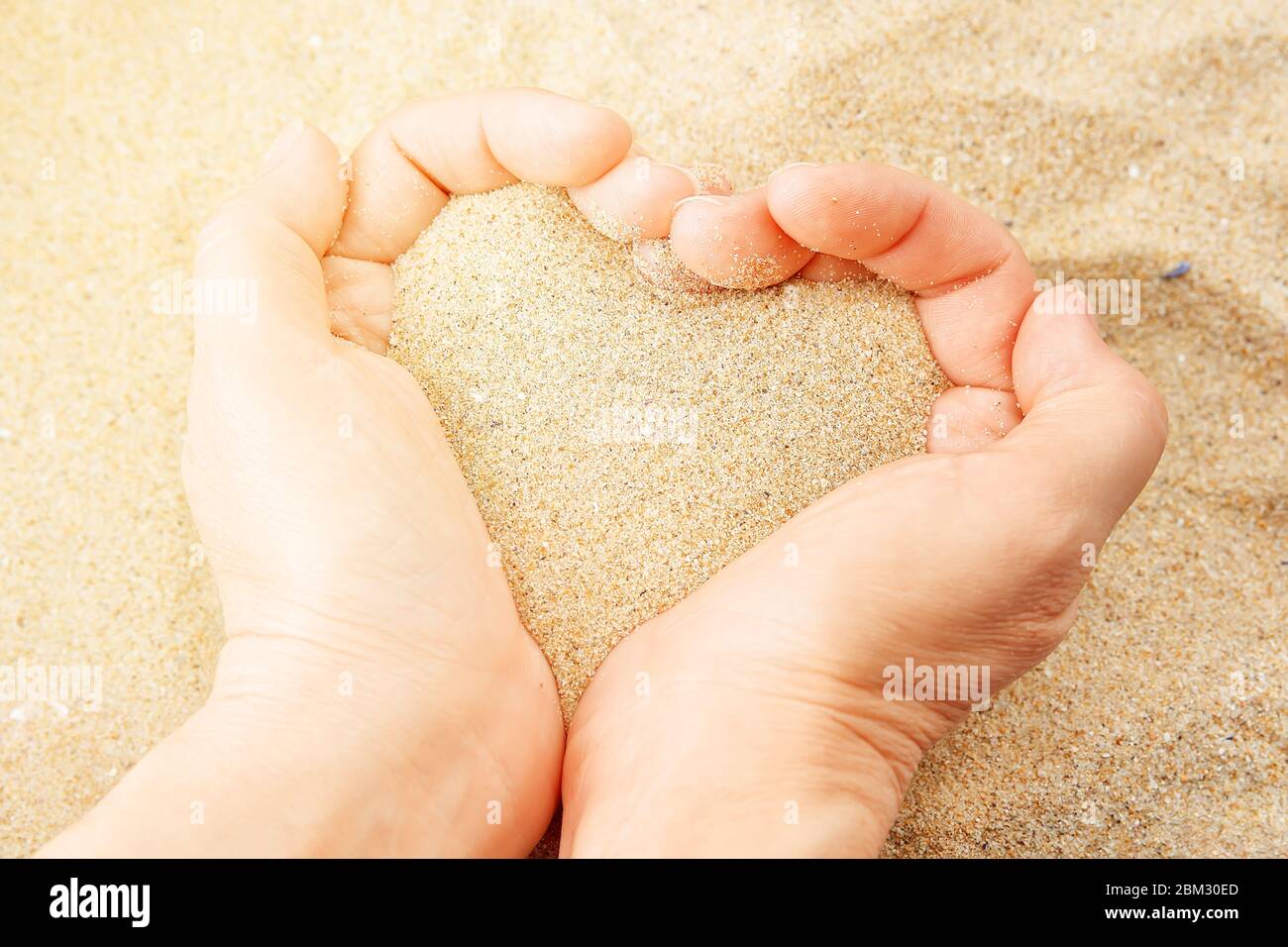 Woman hands hold a heart shaped sea sand in a palm over a beach sand ...
