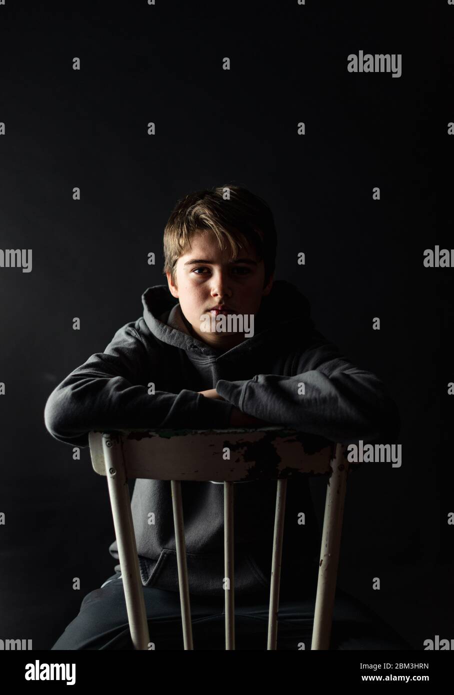 Low key portrait of adolescent boy sitting on a chair in a dark room ...