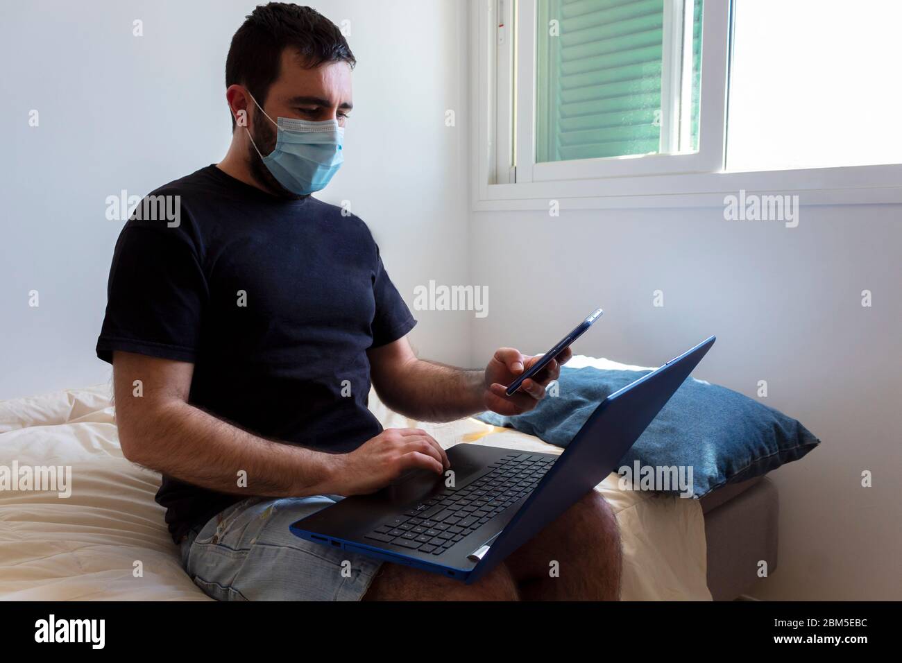 Man teleworking from a room siting on his bed, with an outside window ...