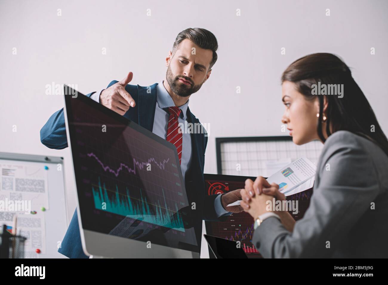 Selective focus of data analyst with papers pointing on graphs on computer monitor to colleague ...