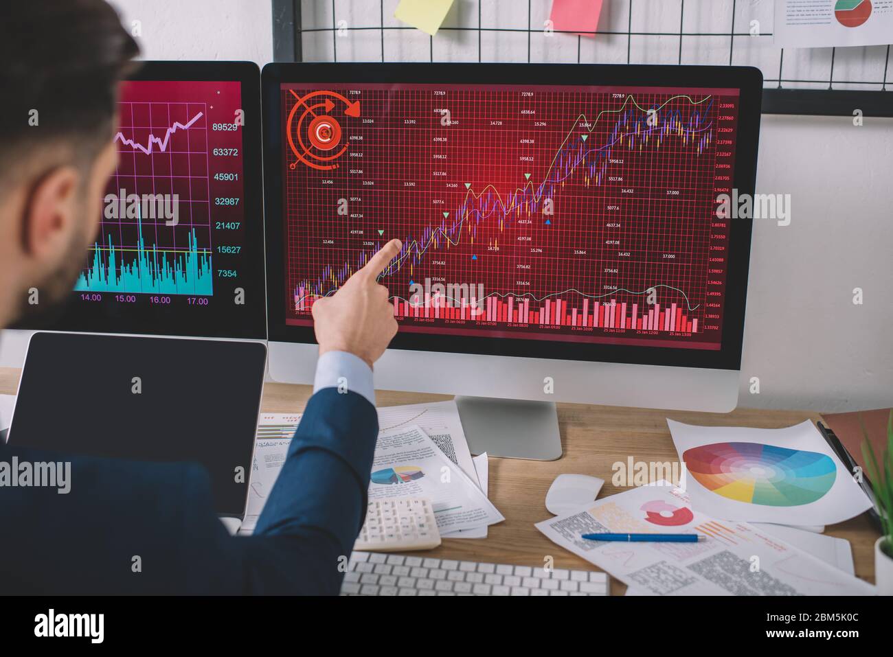 Back view of data analyst pointing on charts on computer monitor near laptop with blank screen ...
