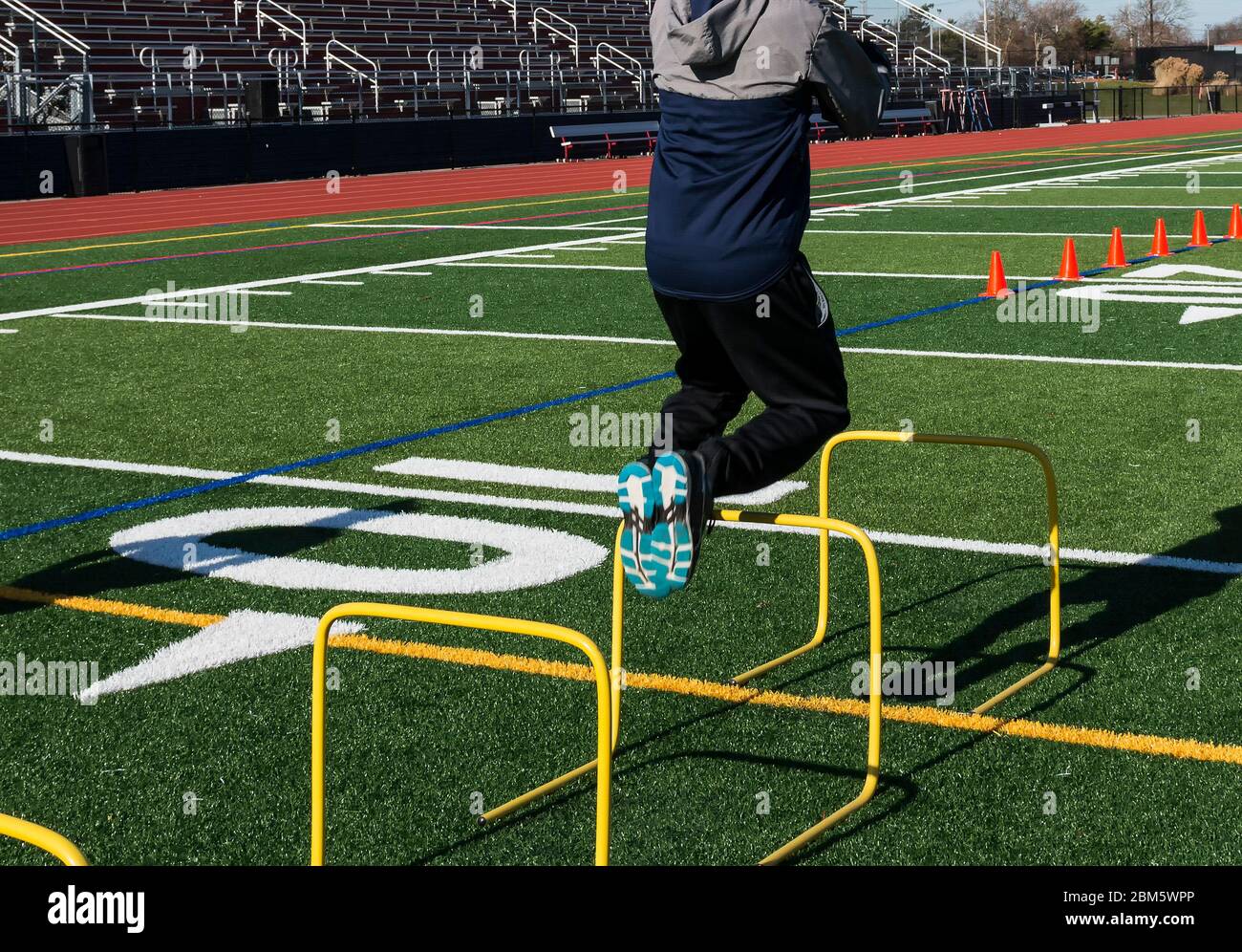A track and field athlete jumping over yellow mini hurdles on a turf field from behind the ...
