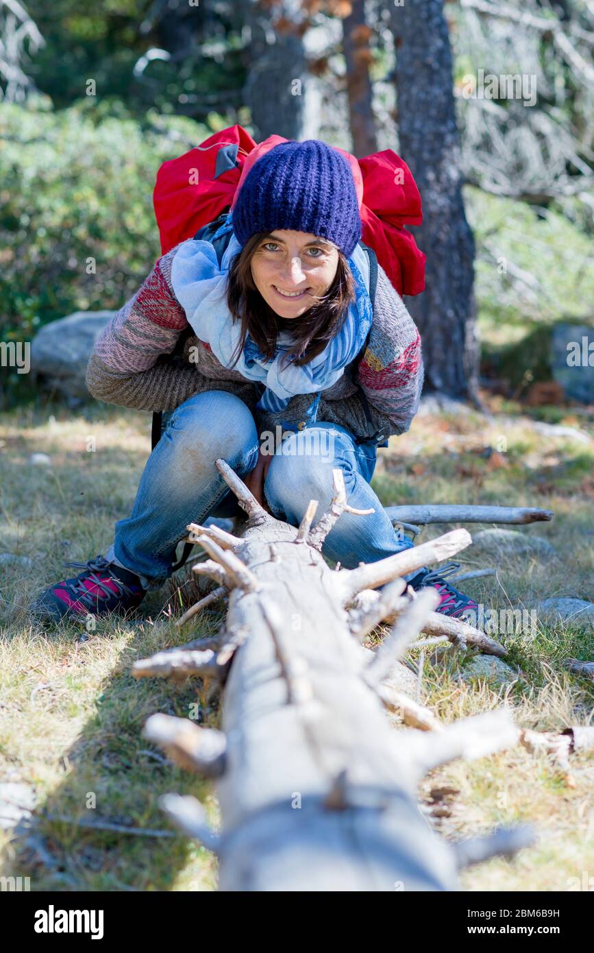 Smiling backpacker woman with wool cap sitting on a log on the ground ...
