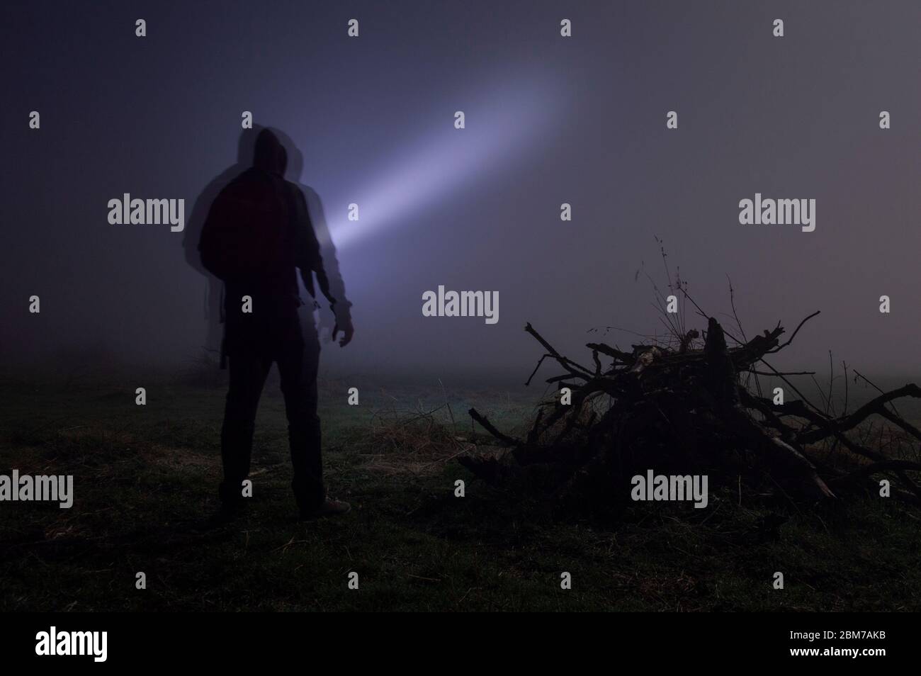 A spooky double exposure of a hooded figure, shining a torch in a foggy ...