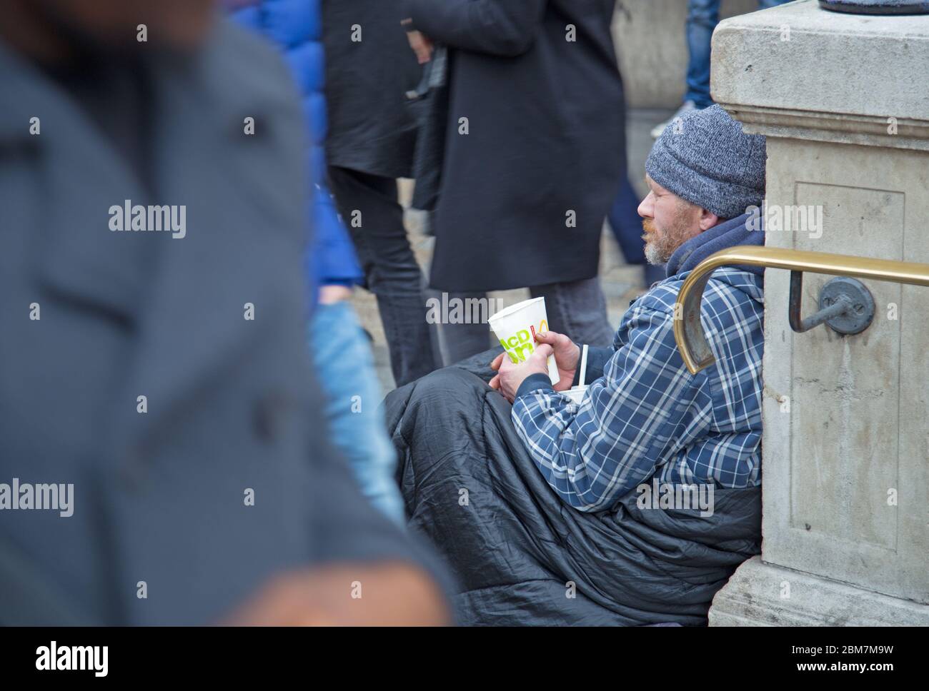 A homeless man sitting in a sleeping bag and holding a cup for ...