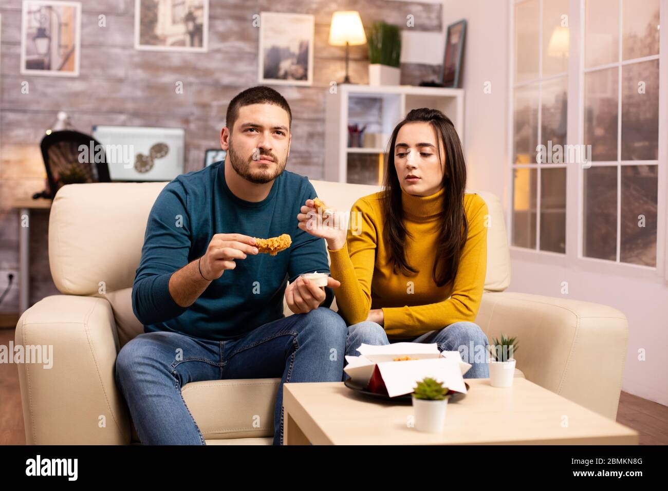 Young couple eating fried chicken in front of the TV in the living room ...