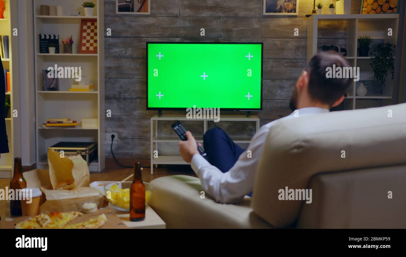 Back view of young man sitting on couch in front of tv with green ...