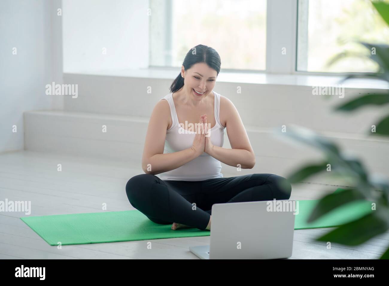 Brunette female performing namaste, having online yoga lesson, smiling ...