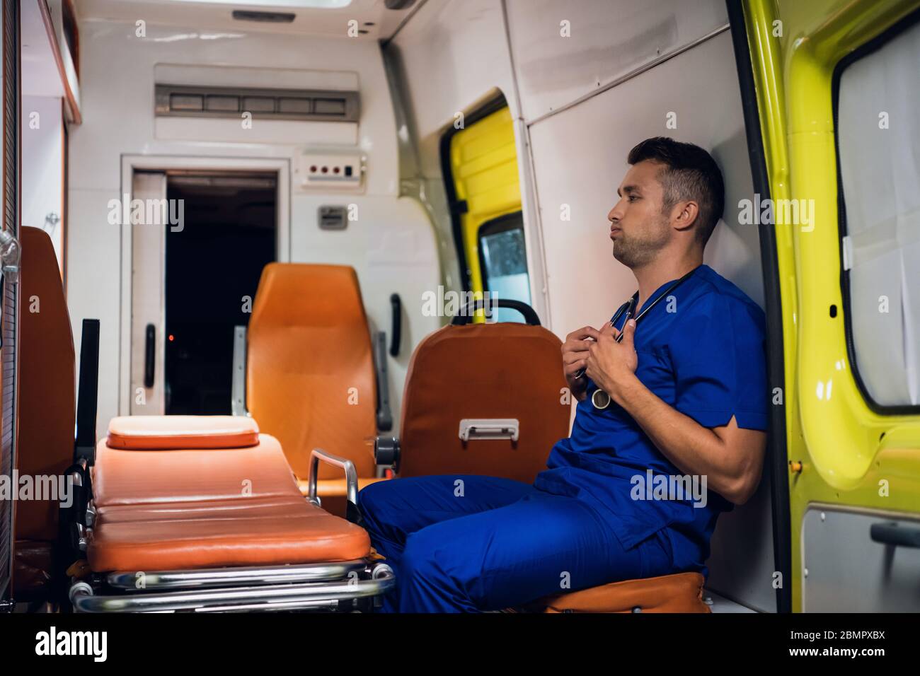Tired paramedic in a blue uniform holding his stethoscope, sitting in ...