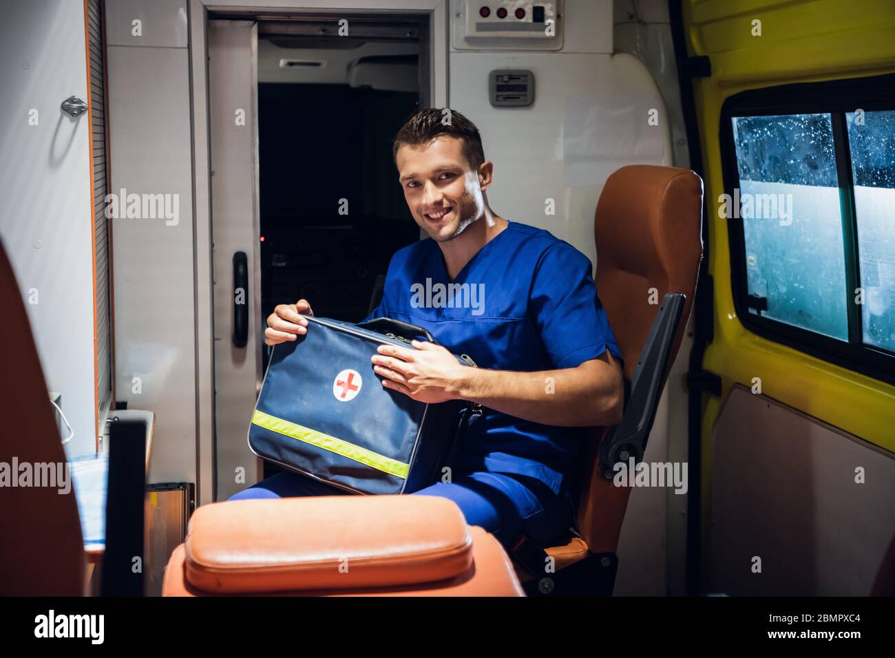 A young male paramedic sitting in an ambulance, holding his medical kit ...