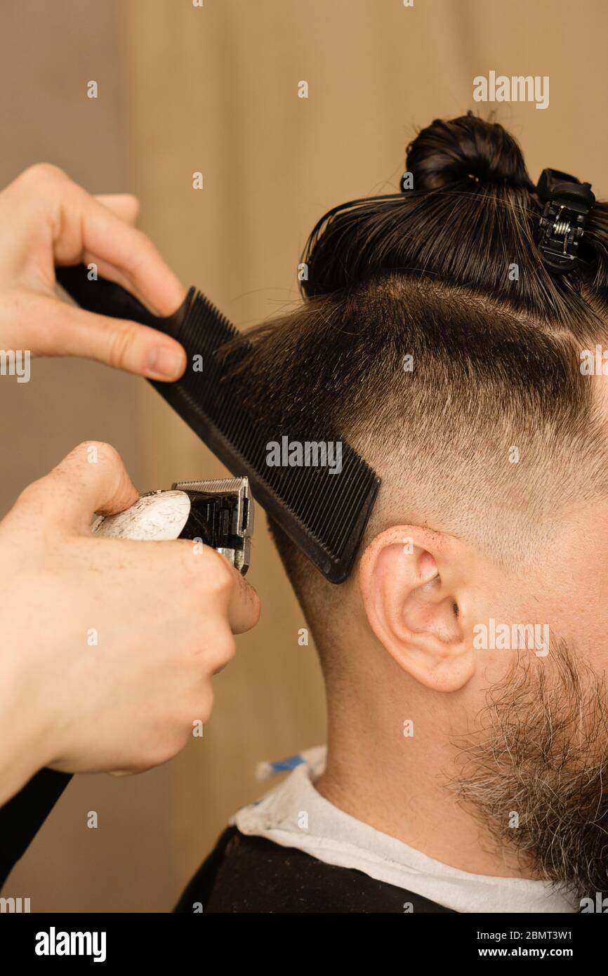Male haircut electric razor. Hand of hairdresser shaves hair with electric shaver. Hipster man getting a trendy haircut with a hair clipper and comb. Barber services. Hairstyling process Stock Photo A...