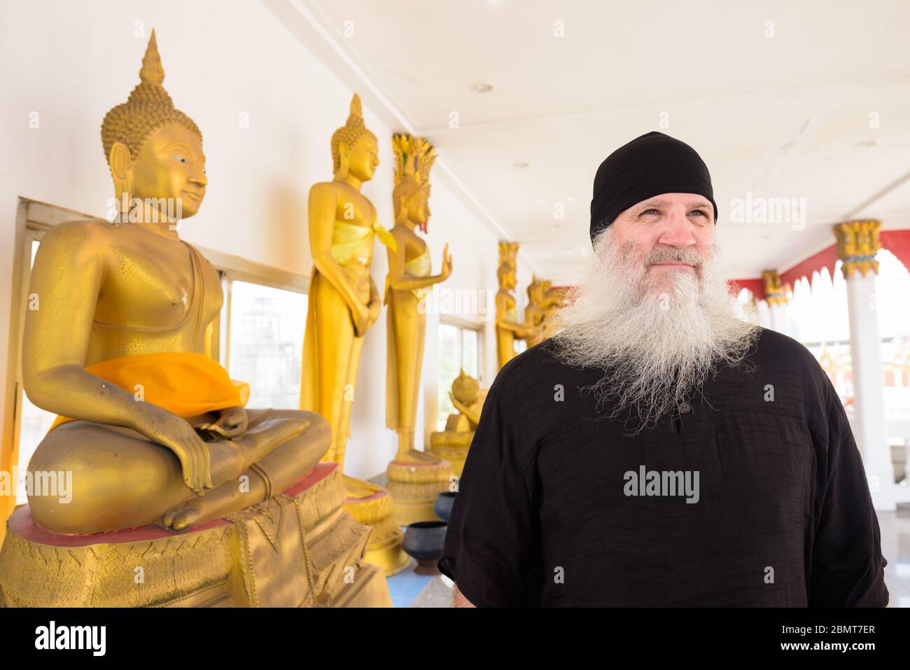 Happy mature bearded tourist man thinking beside golden Buddha statue ...