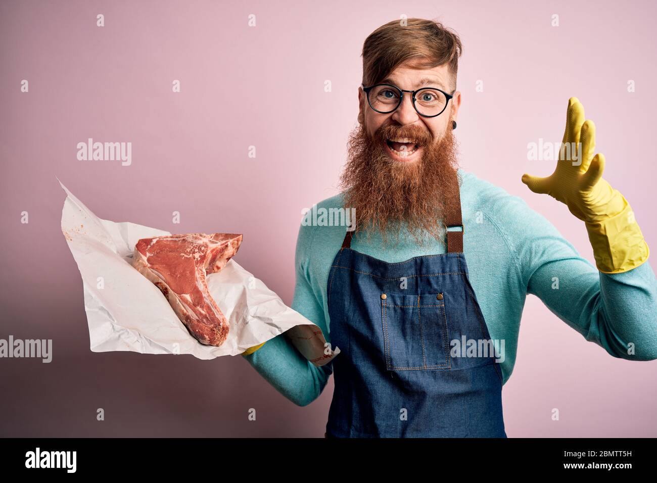 Redhead Irish butcher man with beard holding raw beef steak over pink ...
