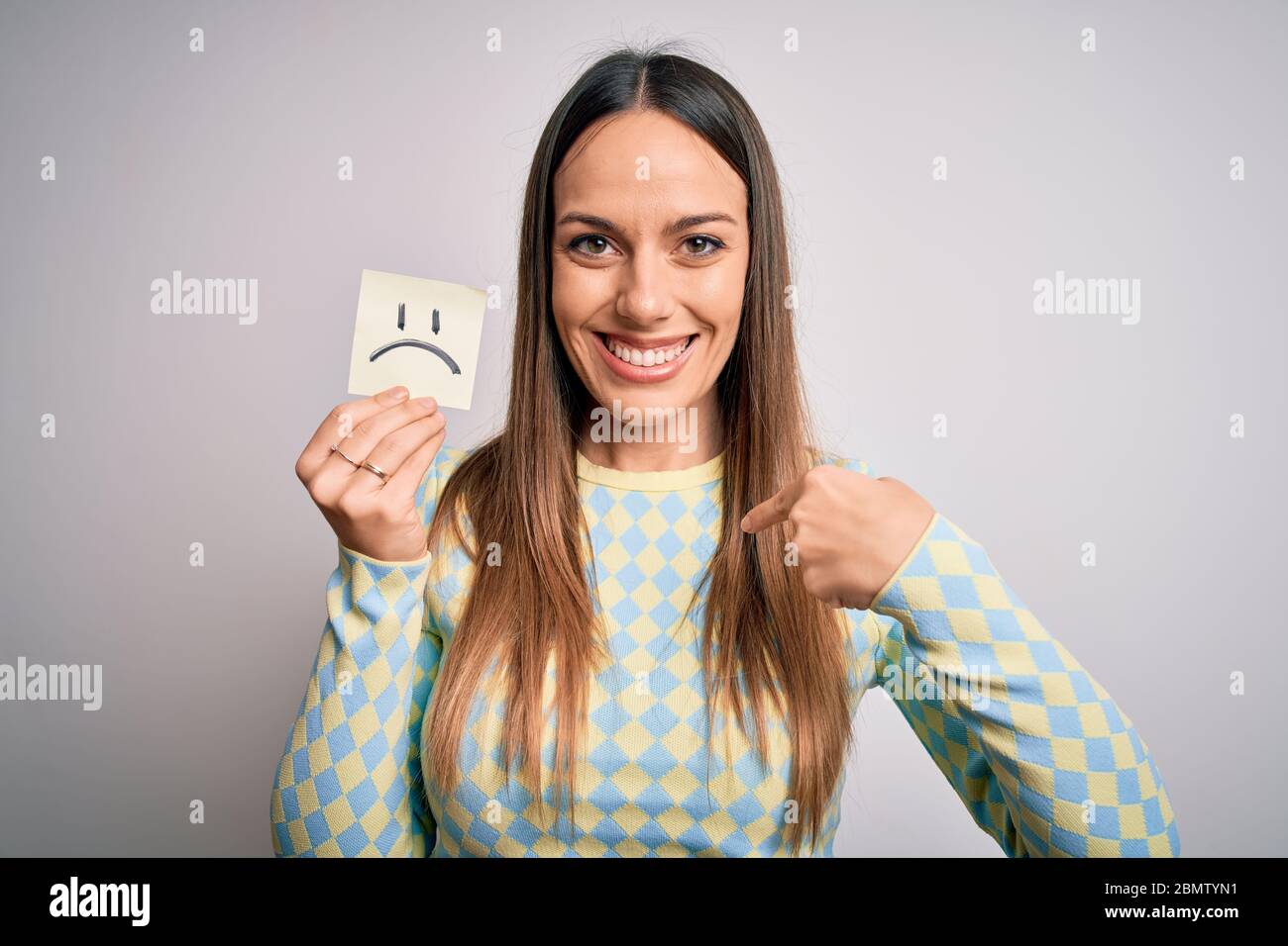 Young blonde woman holding sticky paper note with sad face emoticon ...