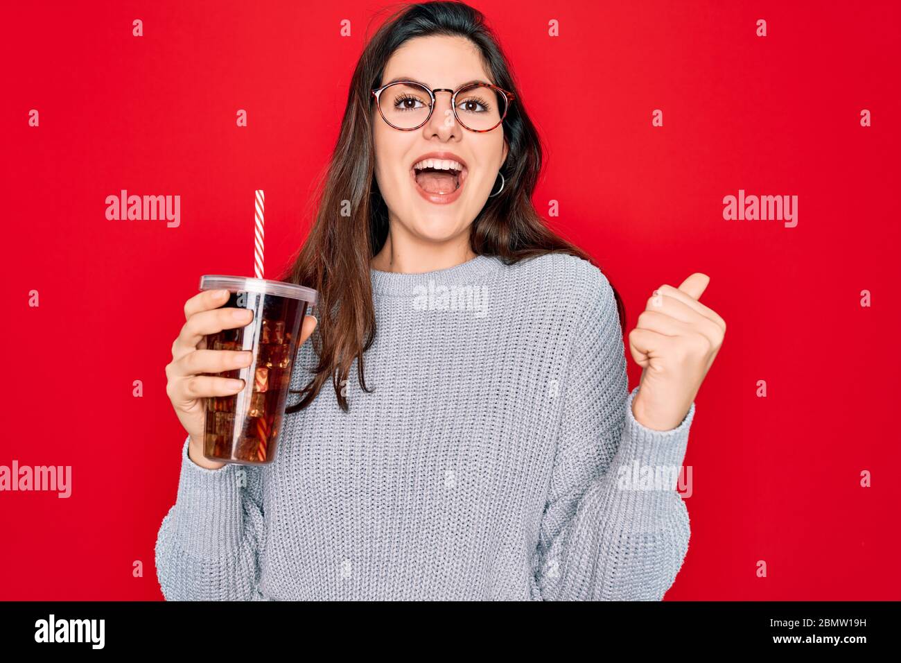 Young beautiful girl wearing glasses drinking sweet drink soda over red ...