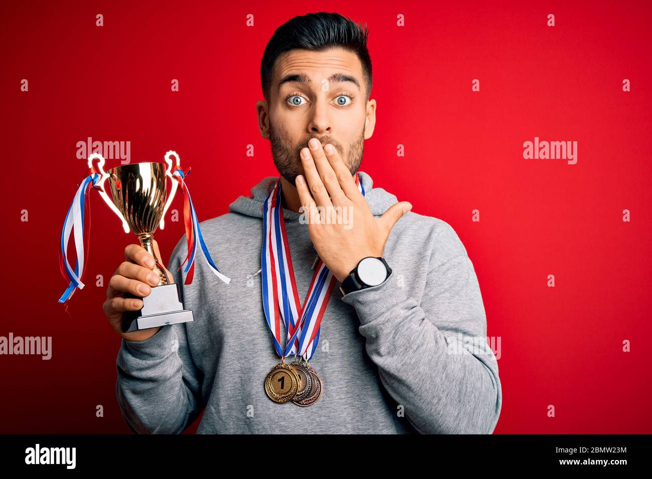 Young handsome succesful man holding trophy wearing medals over red ...