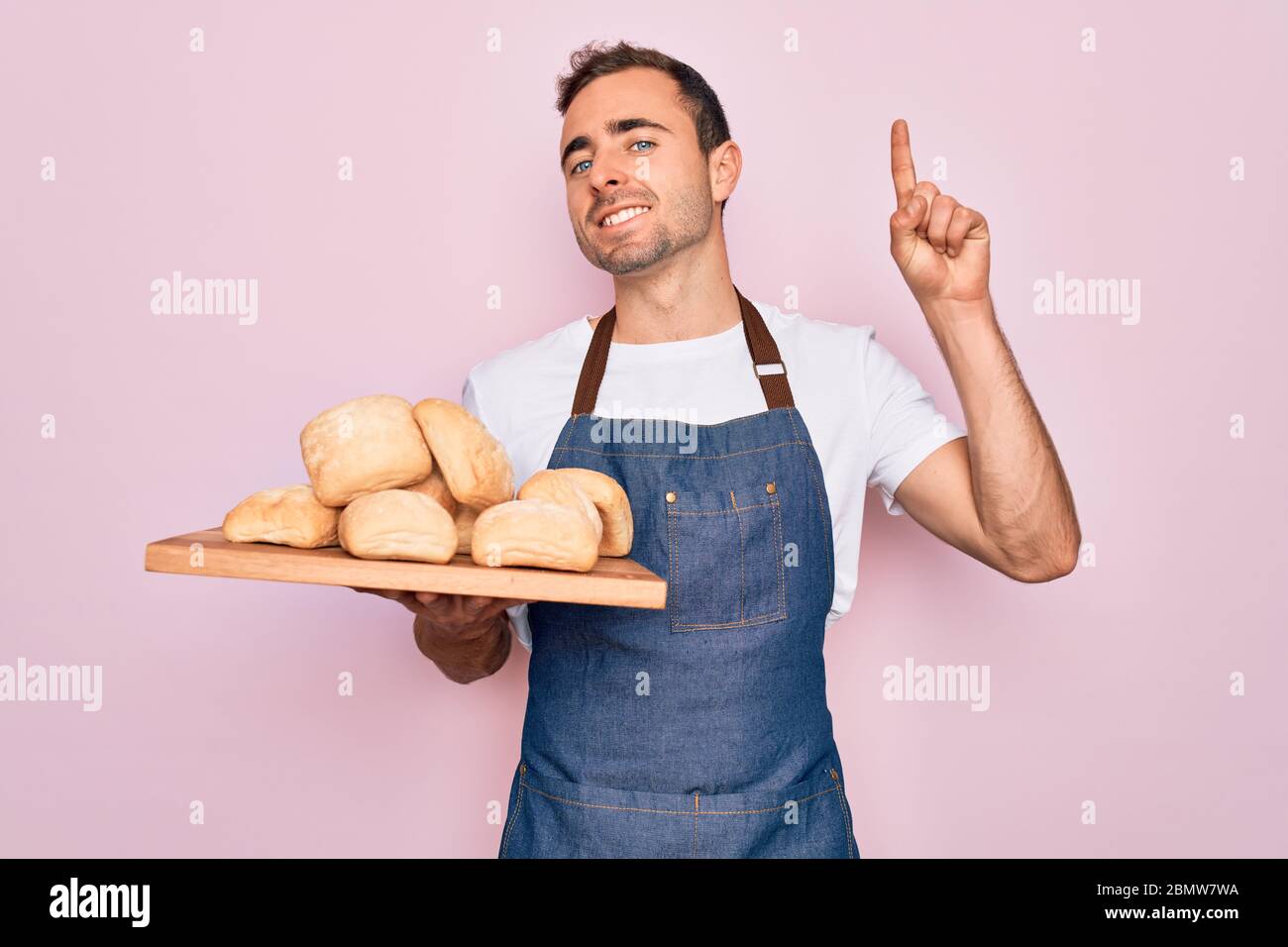 Young handsome baker man with blue eyes wearing apron holding tray with ...