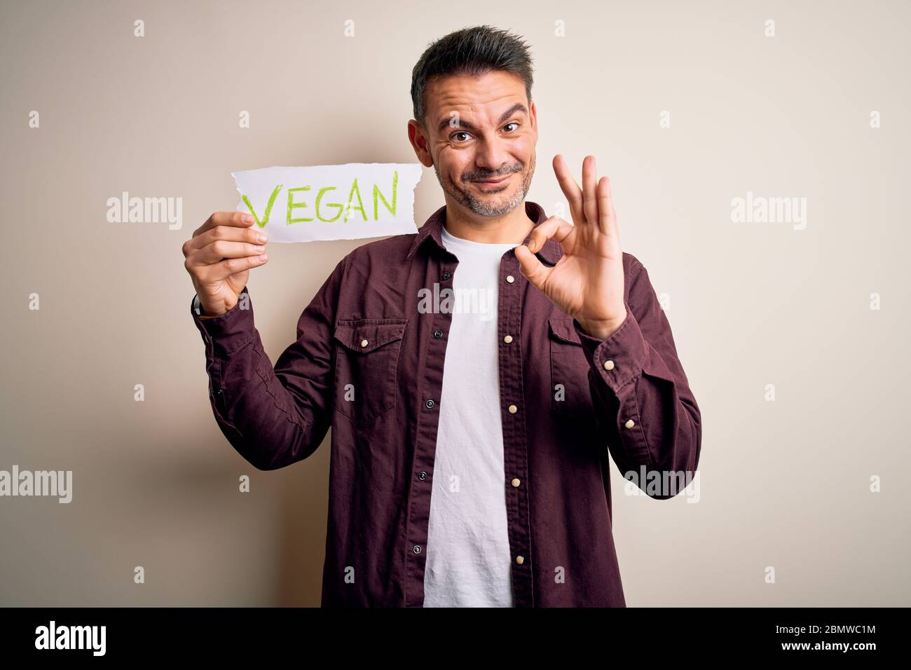 Young handsome veggie man holding paper with vegan message over white ...